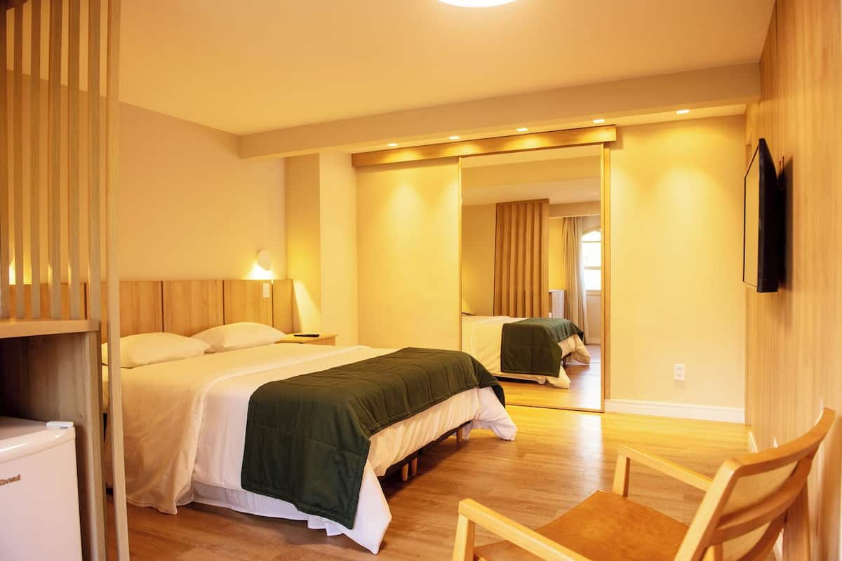Quarto de hotel com cama, mesa de cabeceira, luminária, cadeira e televisão fixada na parede.