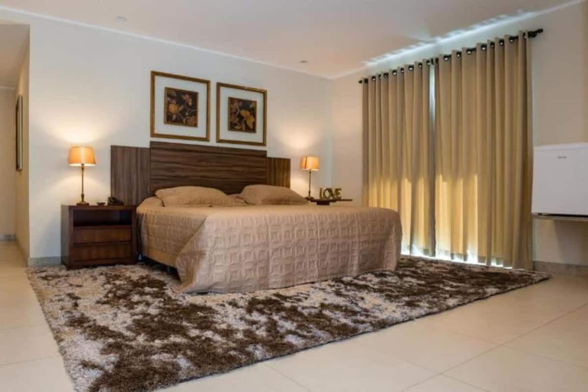 Quarto com cama, duas mesas de cabeceira com luminárias, uma janela grande com cortinas e um refrigerador.