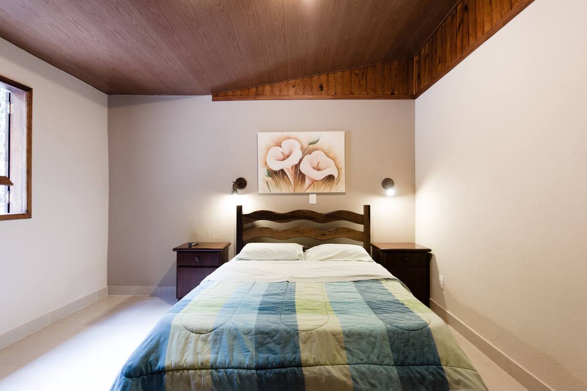 Quarto com cabeceira de madeira, cama com colcha, duas mesas de cabeceira e um quadro na parede.