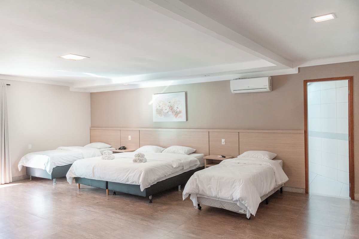 Quarto de hotel com três camas, piso de madeira, luminária no teto e ar-condicionado fixado na parede.