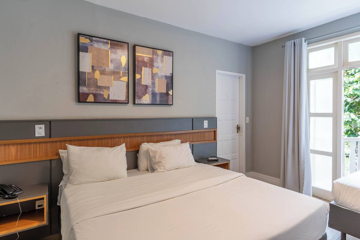 Quarto de hotel com uma cama grande, mesas de cabeceira, um criado-mudo, telefone e duas pinturas abstratas na parede.