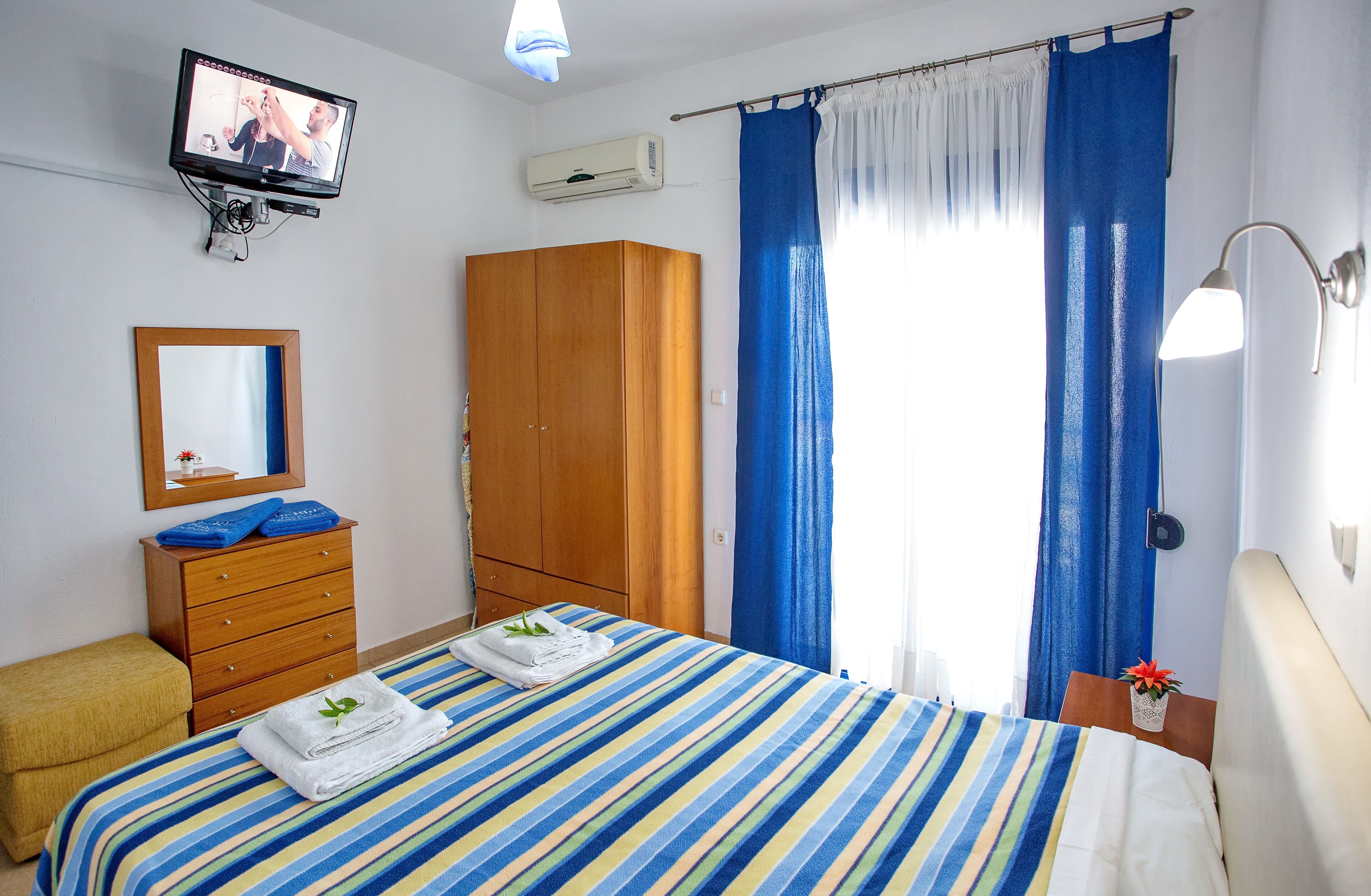 Quarto de hotel com cama, guarda-roupa de madeira, televisão na parede e janela com cortinas azuis.