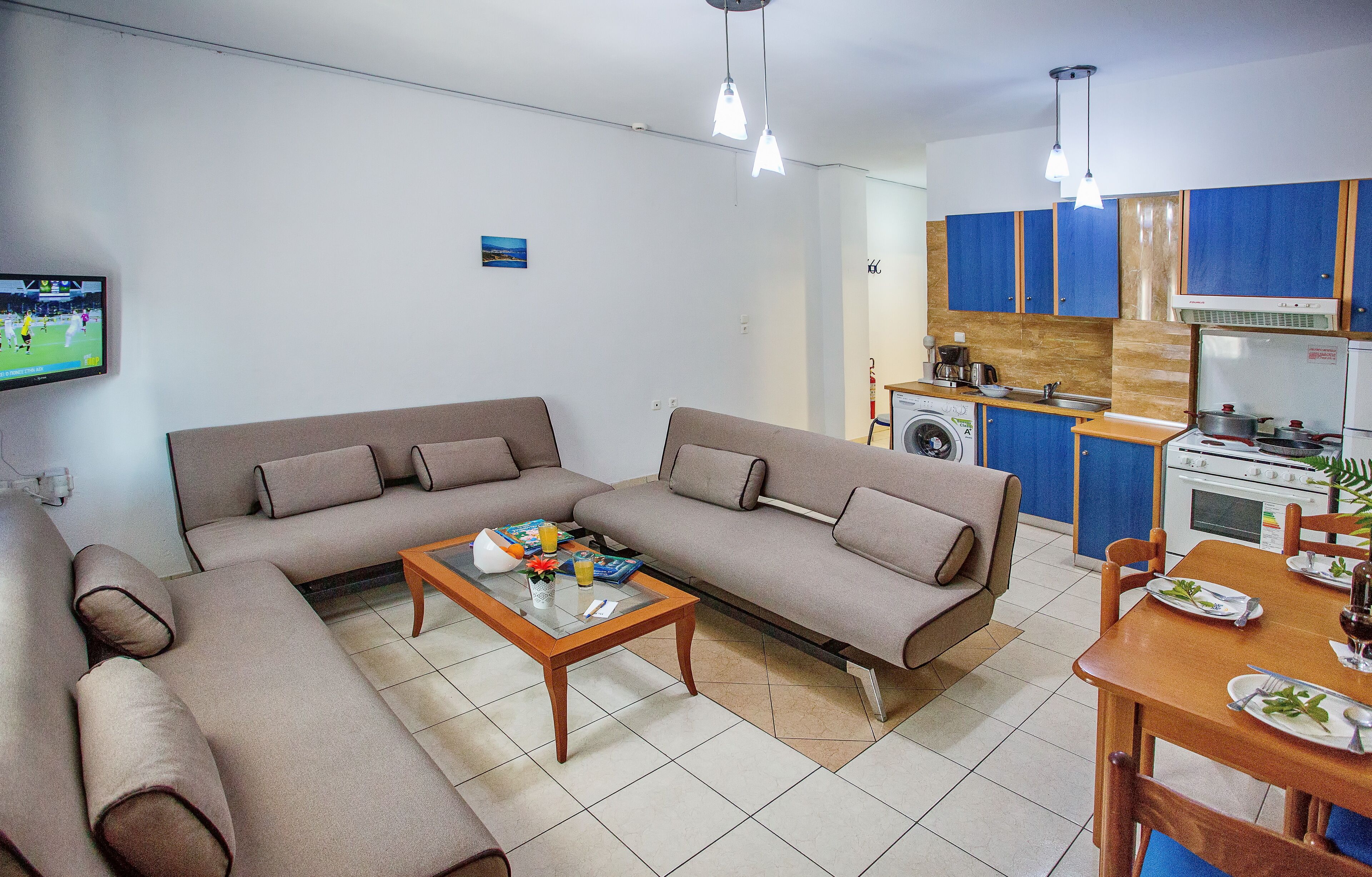 Uma sala moderna com um sofá seccional, uma mesa de centro e um espaço de jantar com mesa posta para dois. Uma cozinha com armários azuis e uma máquina de lavar roupa é visível ao fundo.