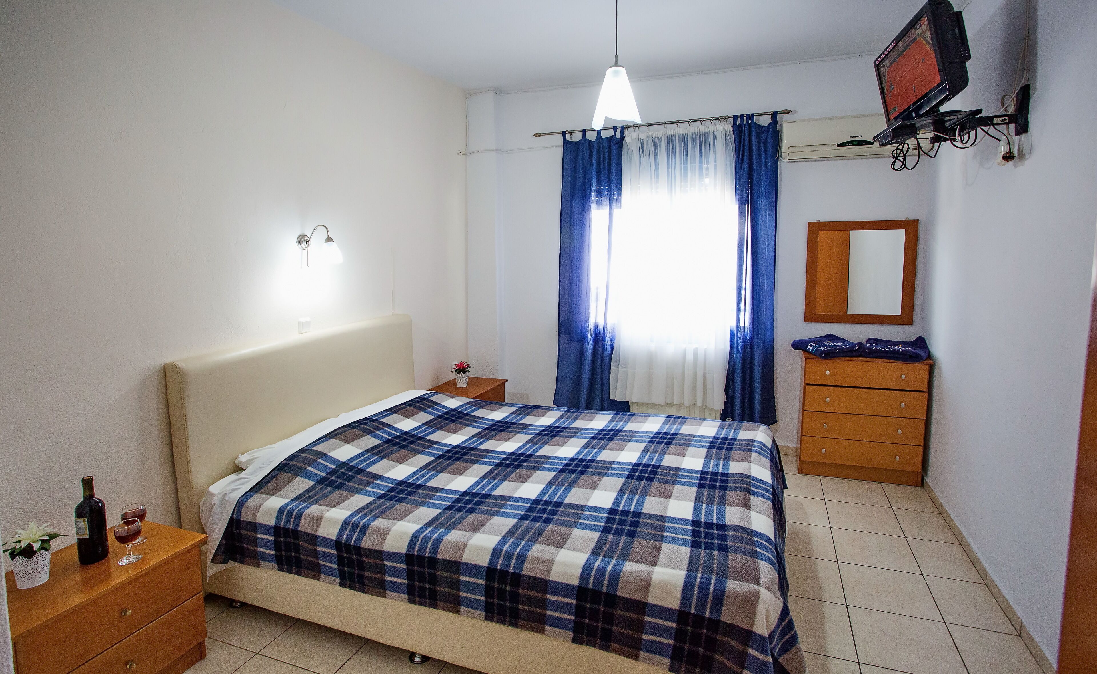 Quarto com cama, penteadeira de madeira, mesa de cabeceira, televisão fixada na parede e janela com cortinas.