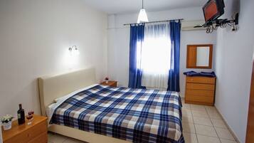 Quarto com cama, penteadeira de madeira, mesa de cabeceira, televisão fixada na parede e janela com cortinas.