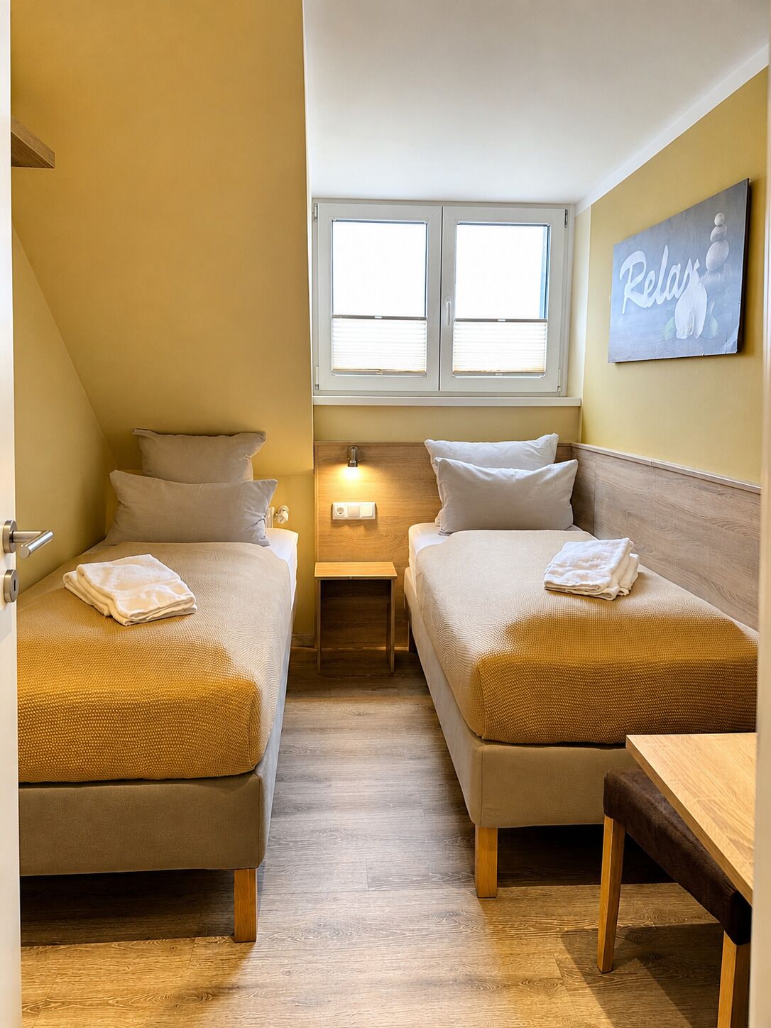 Quarto com duas camas, uma mesinha e janela com persianas.