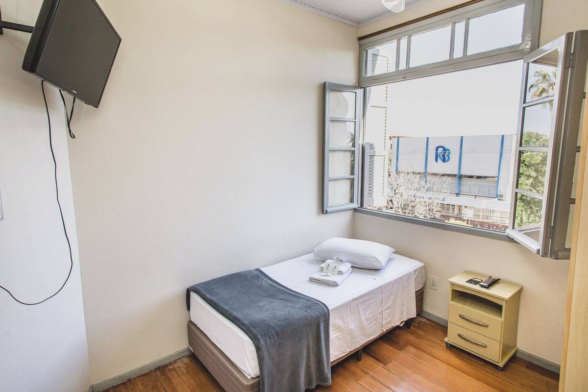 Quarto pequeno com uma cama de solteiro, mesa de cabeceira, televisão na parede e uma janela com persiana aberta.