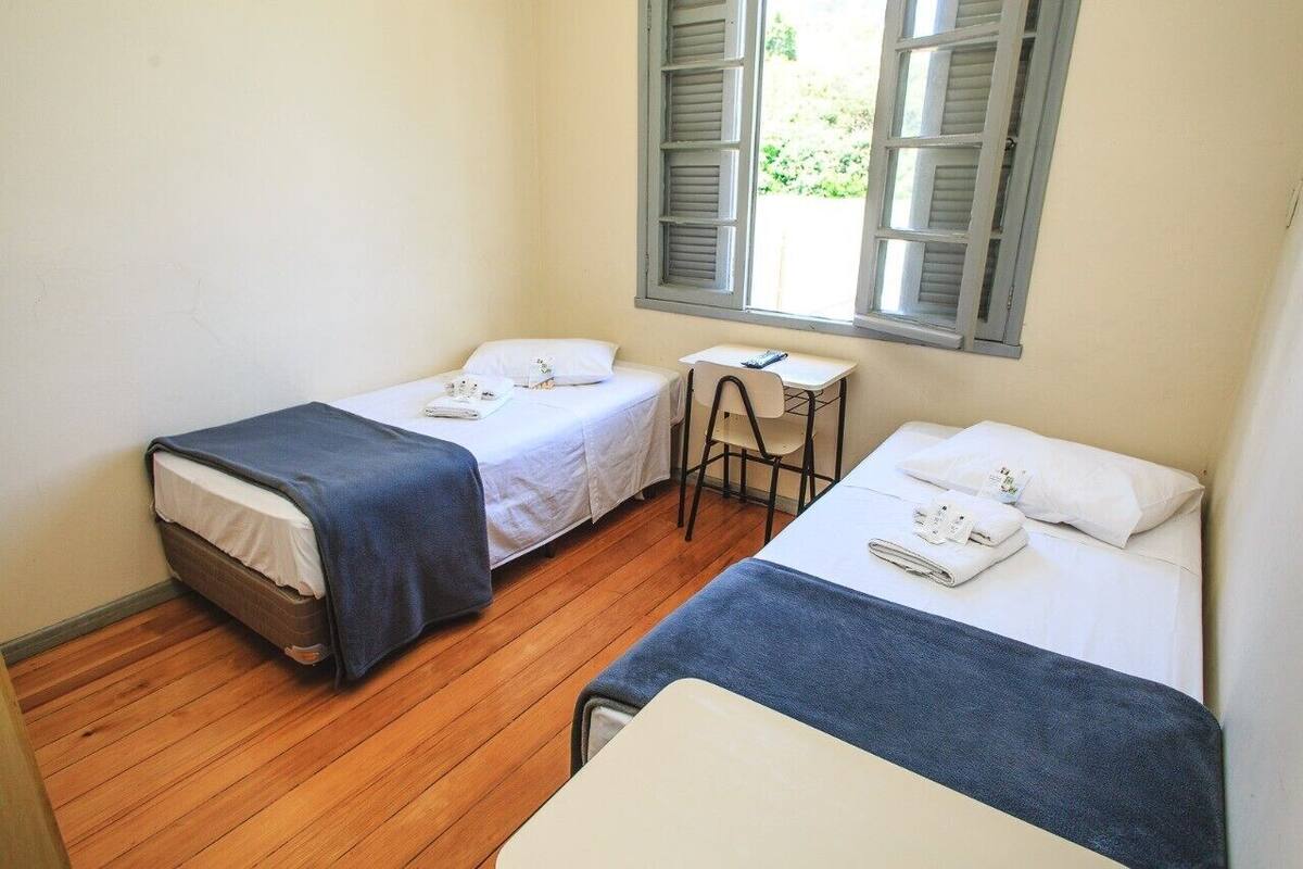 Quarto pequeno com duas camas, uma escrivaninha e uma cadeira.