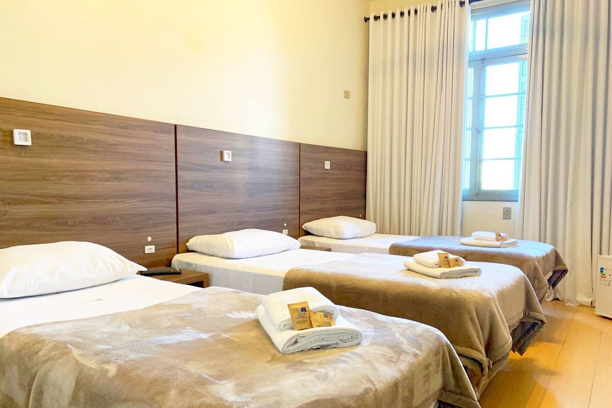 Quarto de hotel com quatro camas de solteiro, cabeceiras de madeira e uma janela grande com cortinas.