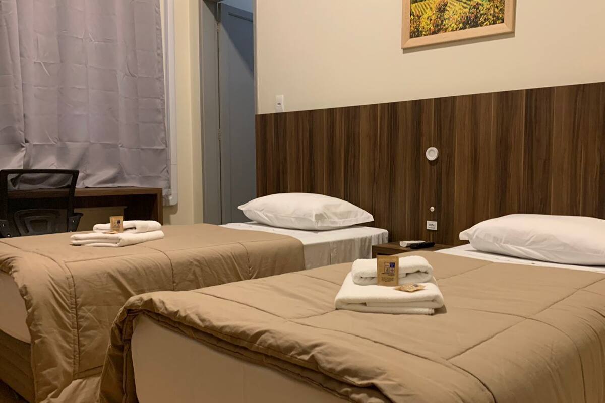 Quarto de hotel com duas camas, cada uma com colcha bege e almofadas brancas. Apresenta parede de painéis de madeira e um pequeno quadro acima das camas.