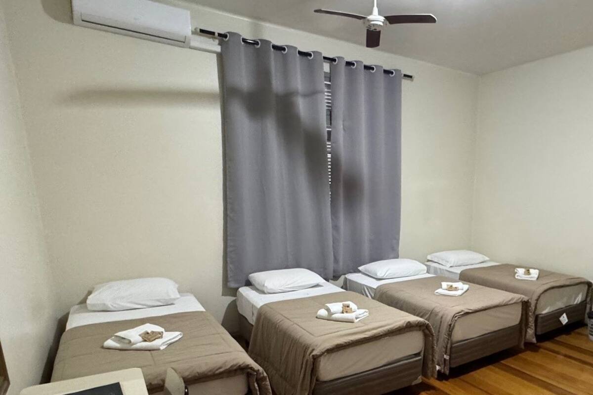 Quarto com três camas de solteiro, cada uma com travesseiro e toalha. Há um ventilador de teto e um aparelho de ar condicionado na parede.