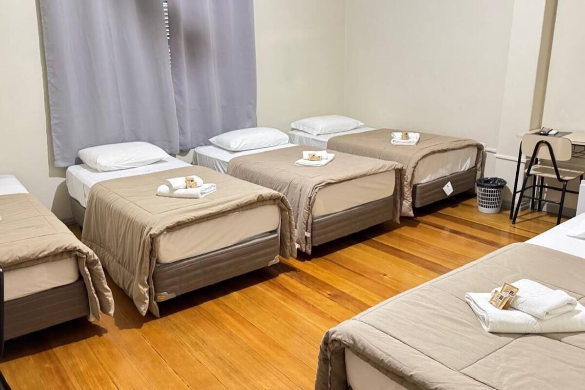Quarto de hotel com quatro camas de solteiro, uma mesinha com uma cadeira, uma janela com cortinas e uma lixeira.