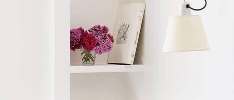Um espaço de trabalho com um laptop sobre uma mesa branca, uma cadeira, um abajur e uma estante com flores e um porta-retrato.