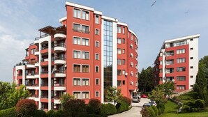 Um edifício residencial de vários andares com varandas, um eixo de elevador de vidro central e um jardim bem cuidado ao redor.