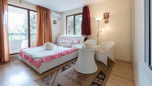 Quarto com cama, mesa redonda e uma cadeira. Há uma janela com cortinas, ar condicionado fixado na parede e um tapete pequeno no chão.