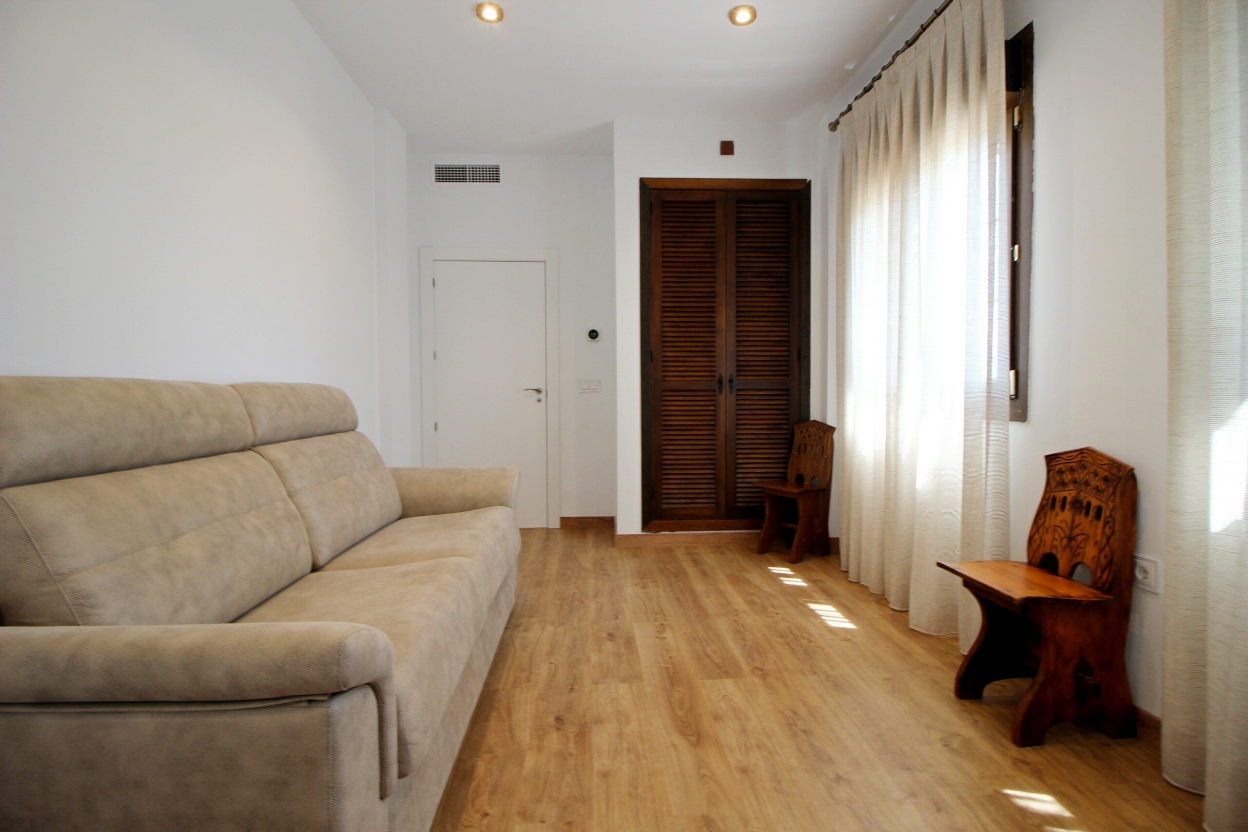 Quarto com sofá bege, piso de madeira, porta de madeira e uma pequena mesa de madeira.