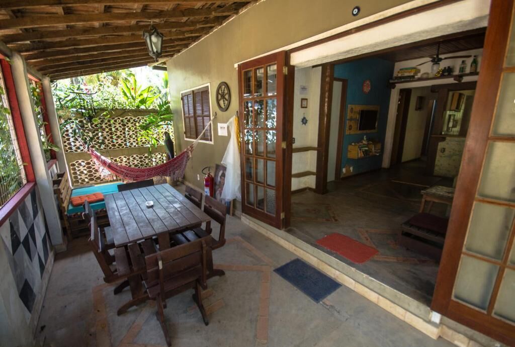 Alpendre coberto com mesa e cadeiras de madeira, uma rede e vista para o jardim.