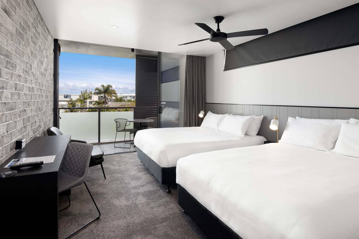 Quarto de hotel moderno com duas camas, uma escrivaninha, uma cadeira, ventoinha no teto e varanda com vista.