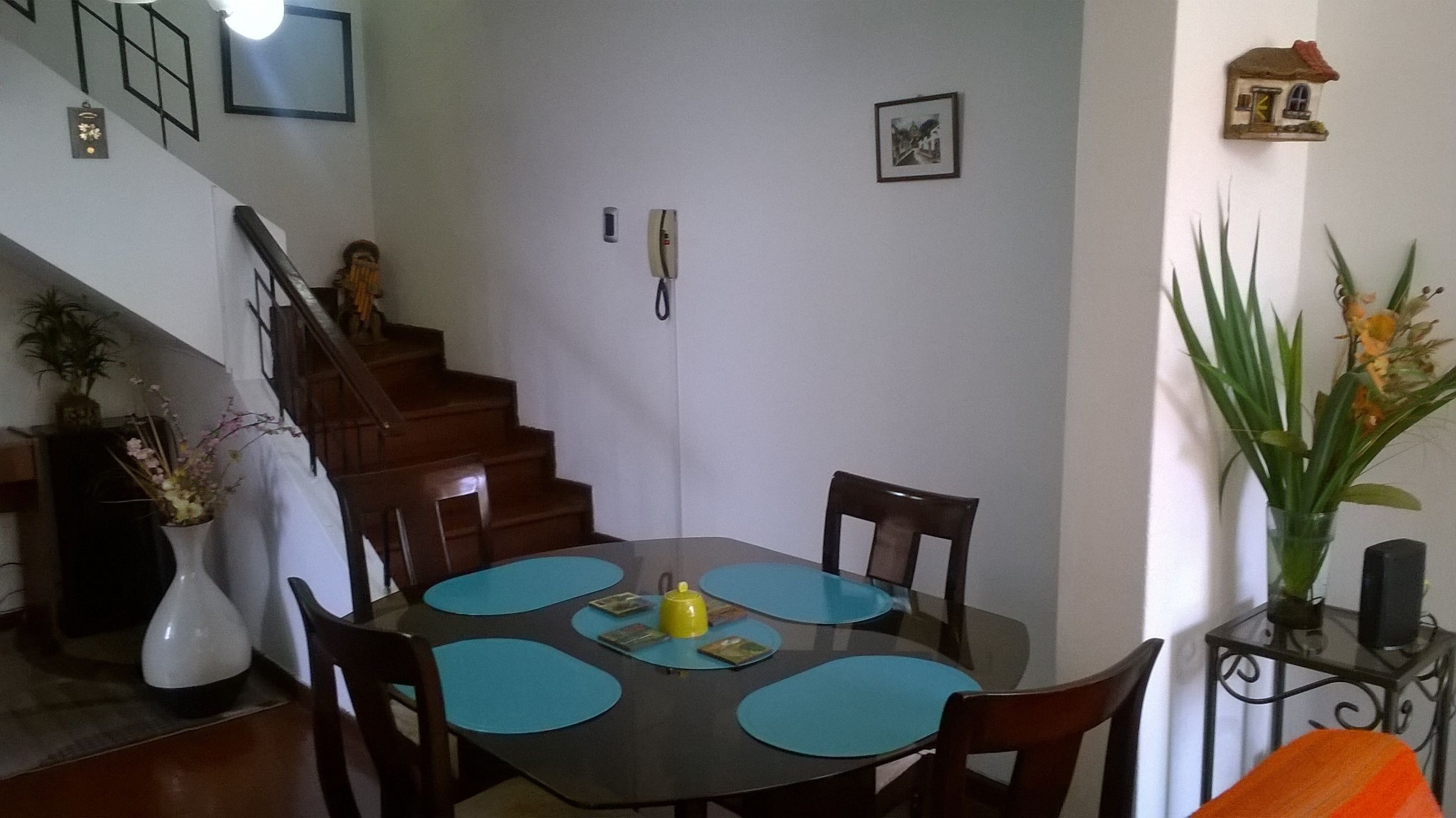 Uma área de refeições com mesa, cadeiras, uma escada e uma pequena planta em uma mesinha lateral.