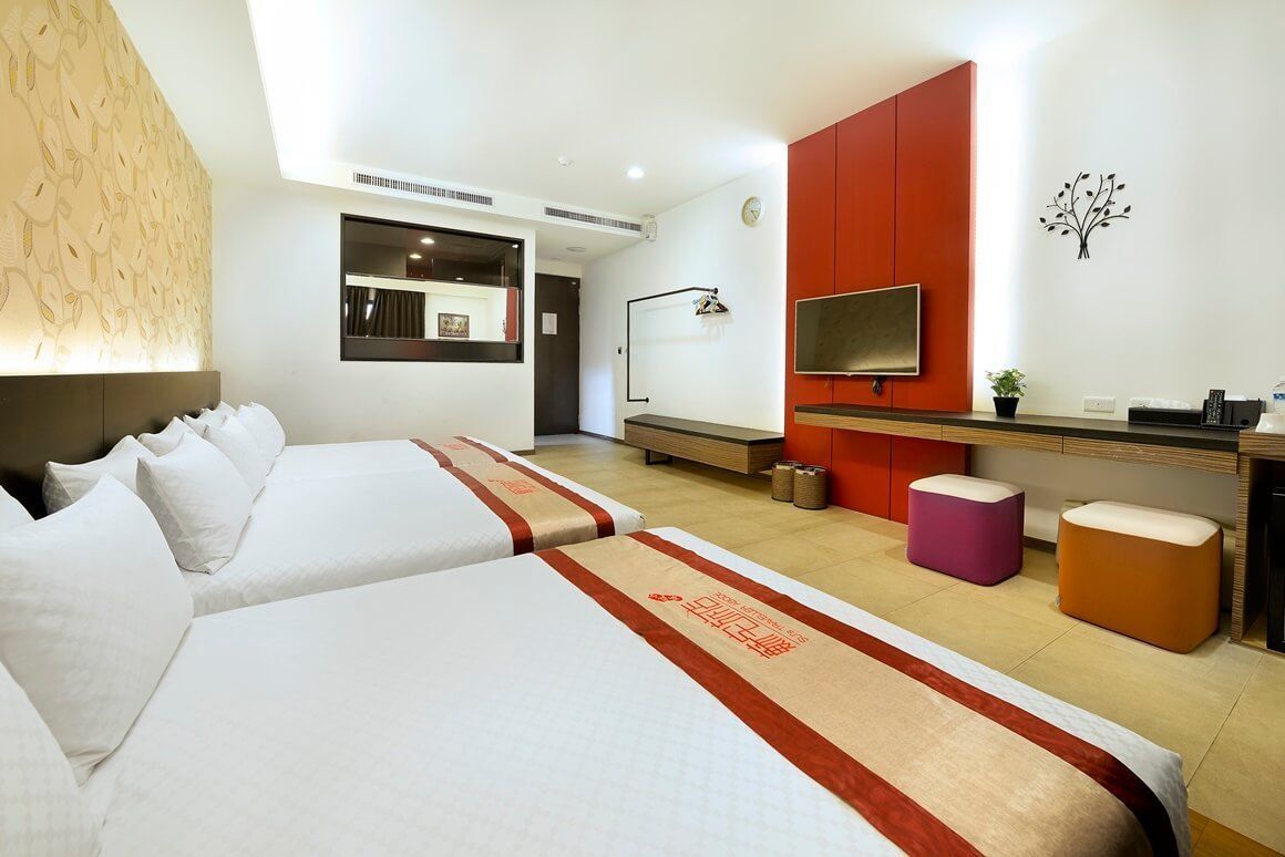 Quarto de hotel com duas camas, uma escrivaninha, uma TV e uma parede em vermelho.