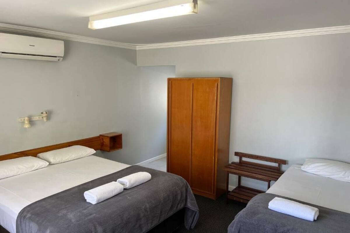 Quarto de hotel com duas camas, um guarda-roupa de madeira, um banco e um aparelho de ar condicionado.