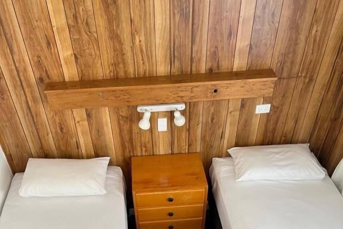 Quarto com duas camas de solteiro, uma mesinha de cabeceira de madeira entre elas e um cobertor vermelho no chão.