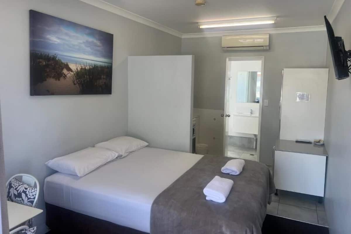 Quarto de hotel com cama, escrivaninha, cadeira, banheiro e televisão na parede.