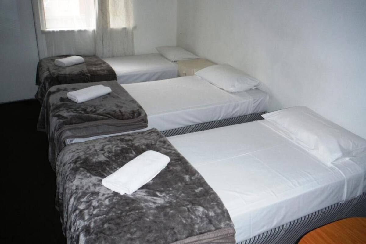 Quarto de hotel com três camas de solteiro, cada uma com uma almofada branca e uma toalha branca dobrada. As camas possuem roupa de cama cinza e branca.
