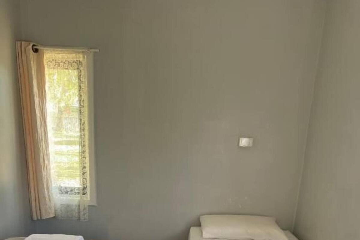 Quarto com duas camas, uma com colcha branca e outra com colcha bege. Há uma janela com cortina e um interruptor na parede.