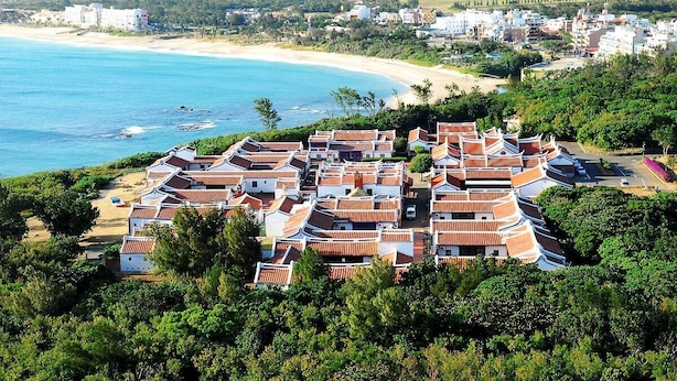 Um resort costeiro com arquitetura tradicional, uma praia e montanhas ao fundo.