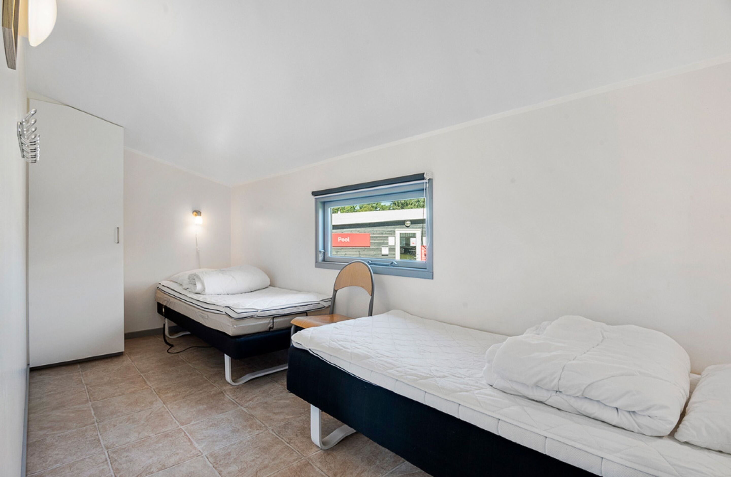 Quarto compacto com uma cama de solteiro, uma escrivaninha pequena e uma janela com vista para uma piscina.