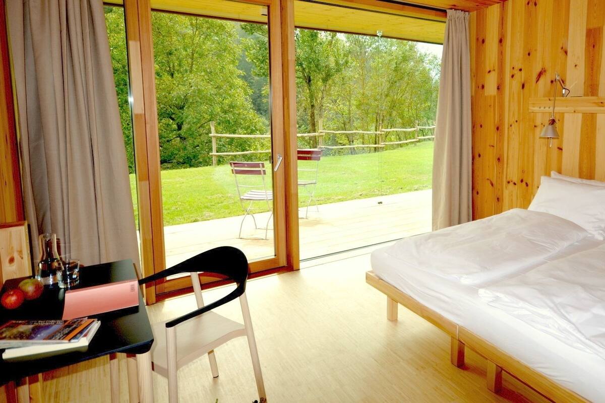 Quarto de madeira com cama, escrivaninha e cadeira. Há uma janela com vista para uma área gramada e árvores.