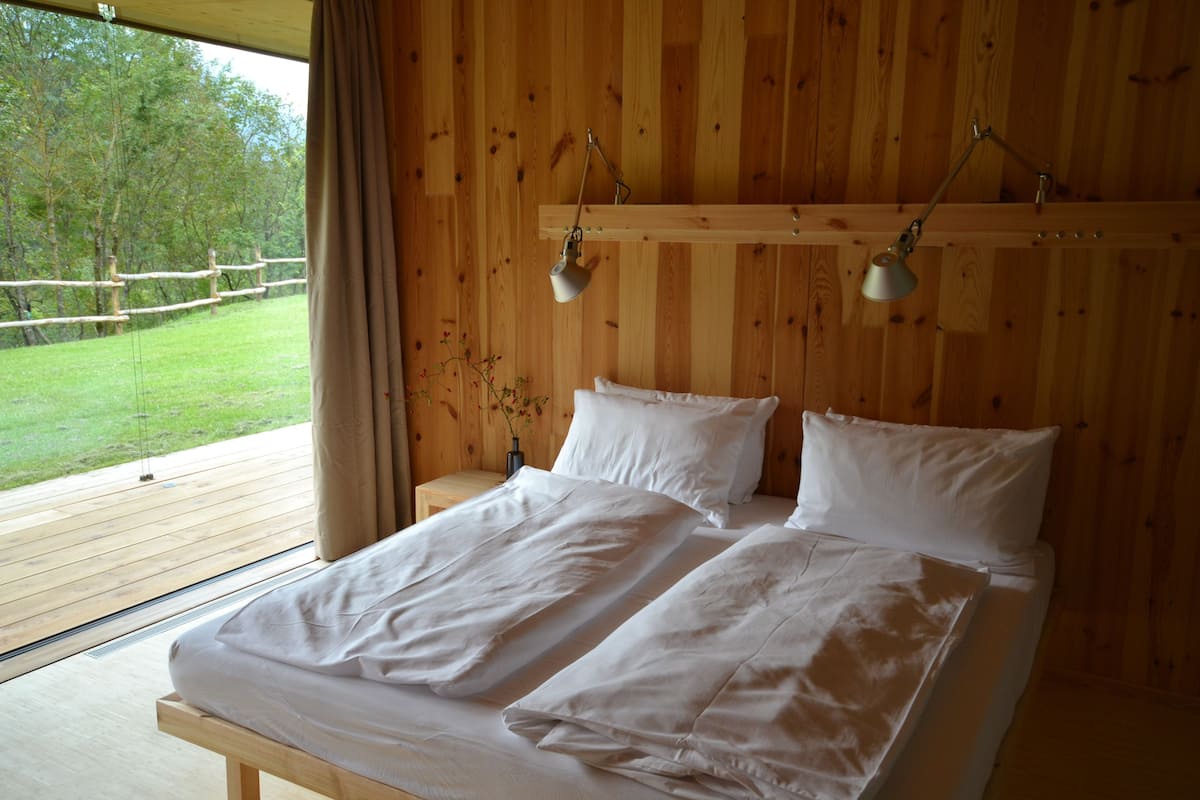 Quarto de madeira com uma cama, duas luminárias de cabeceira e vista para um deck de madeira e vegetação lá fora.