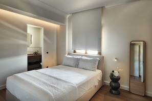 Quarto moderno com uma cama grande, uma mesinha de cabeceira, um espelho e uma luminária fixada na parede.