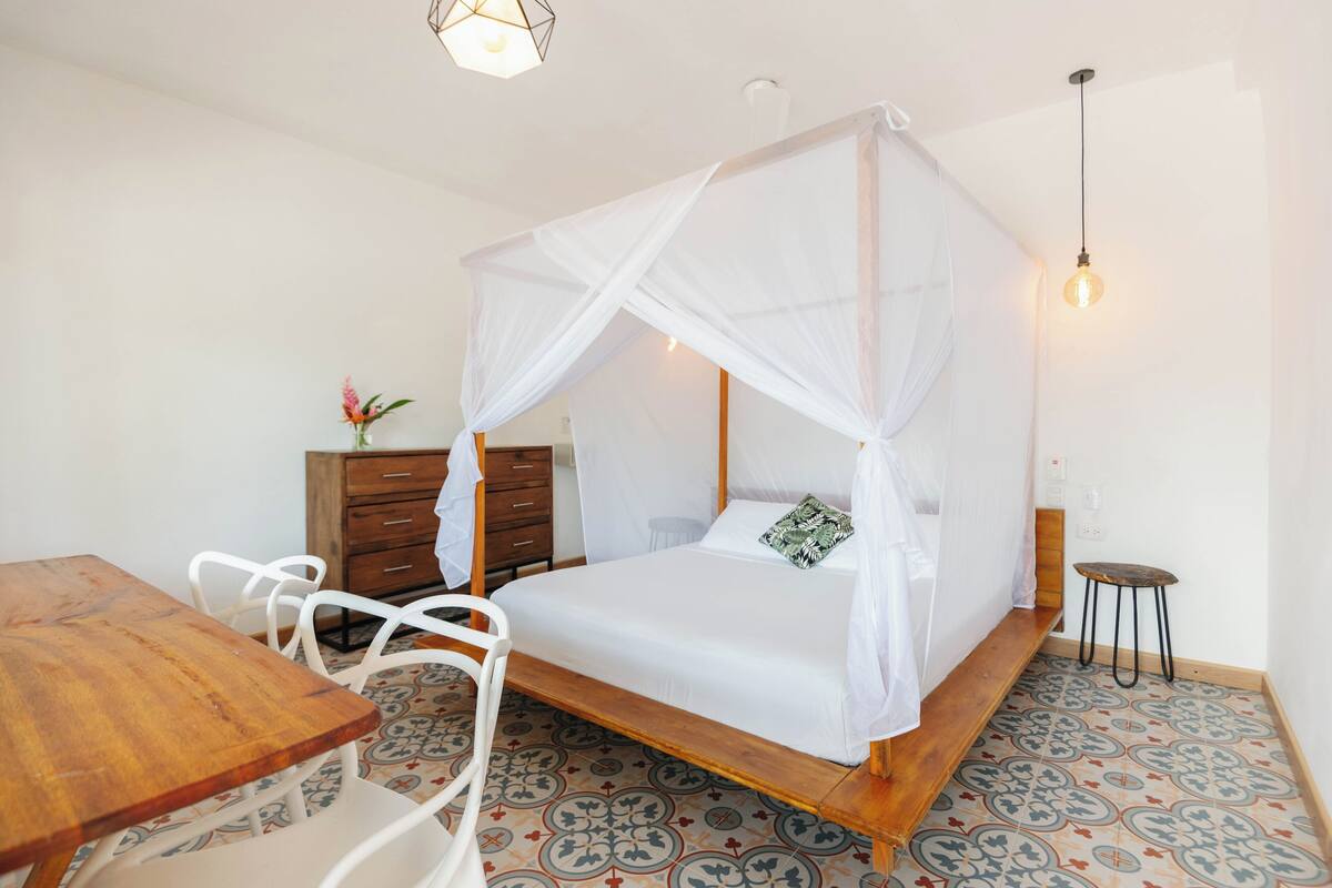 Quarto com cama sob um dossel branco, mesa de madeira com cadeiras, um banquinho pequeno e piso estampado.