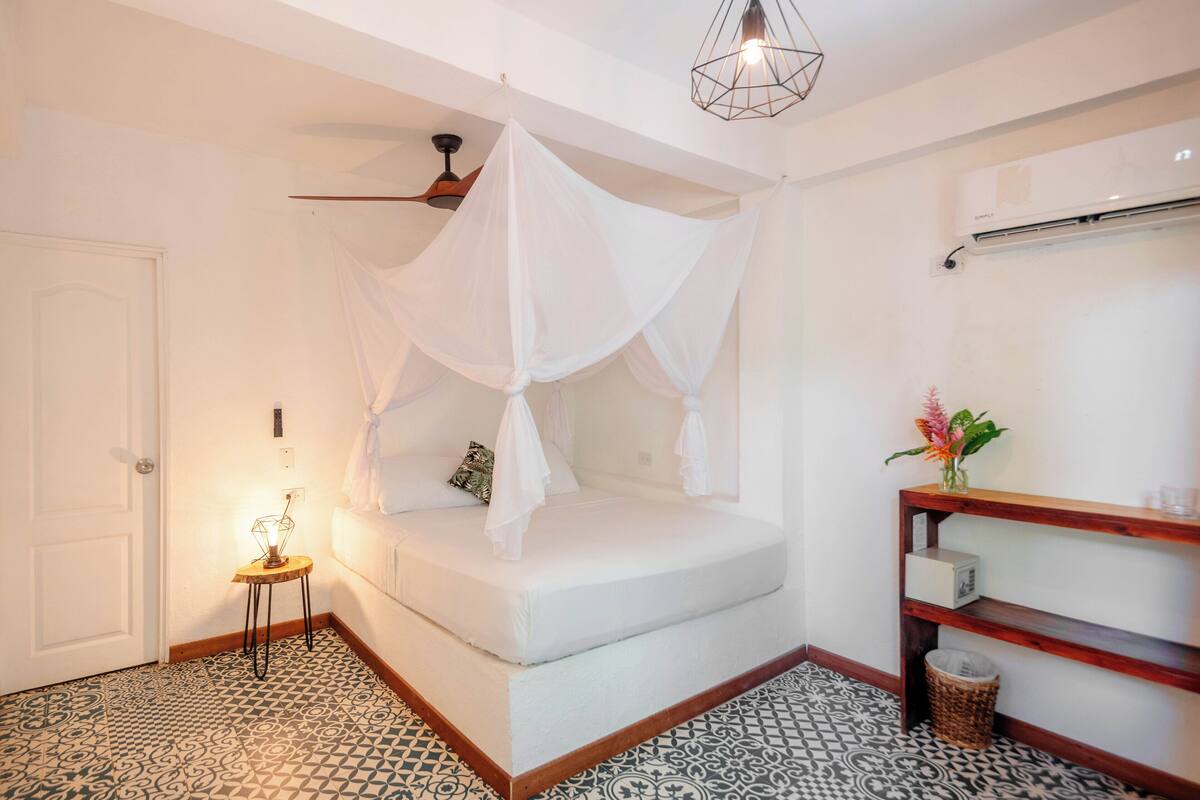 Quarto com cama de dossel, mesinha de cabeceira com abajur, ar-condicionado fixado na parede e prateleira com objetos decorativos.