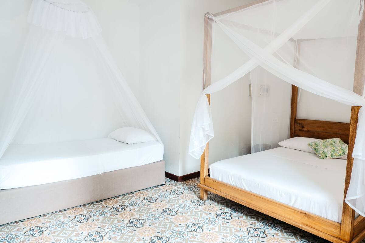 Quarto com uma cama sob um dossel, uma cama menor com um banco, e um piso estampado.