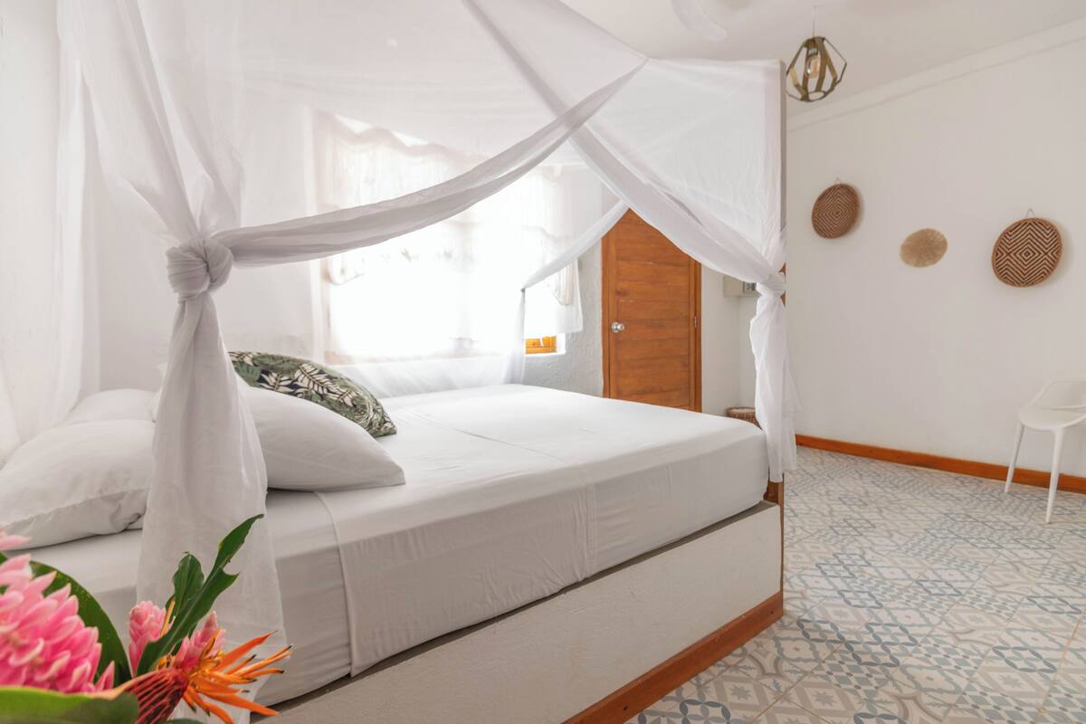 Cama bem arrumada com roupa de cama branca e um dossel, uma porta de madeira e um piso estampado.