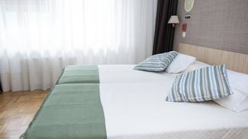 Quarto de hotel com cama, abajur, relógio na parede e janela com cortinas leves.