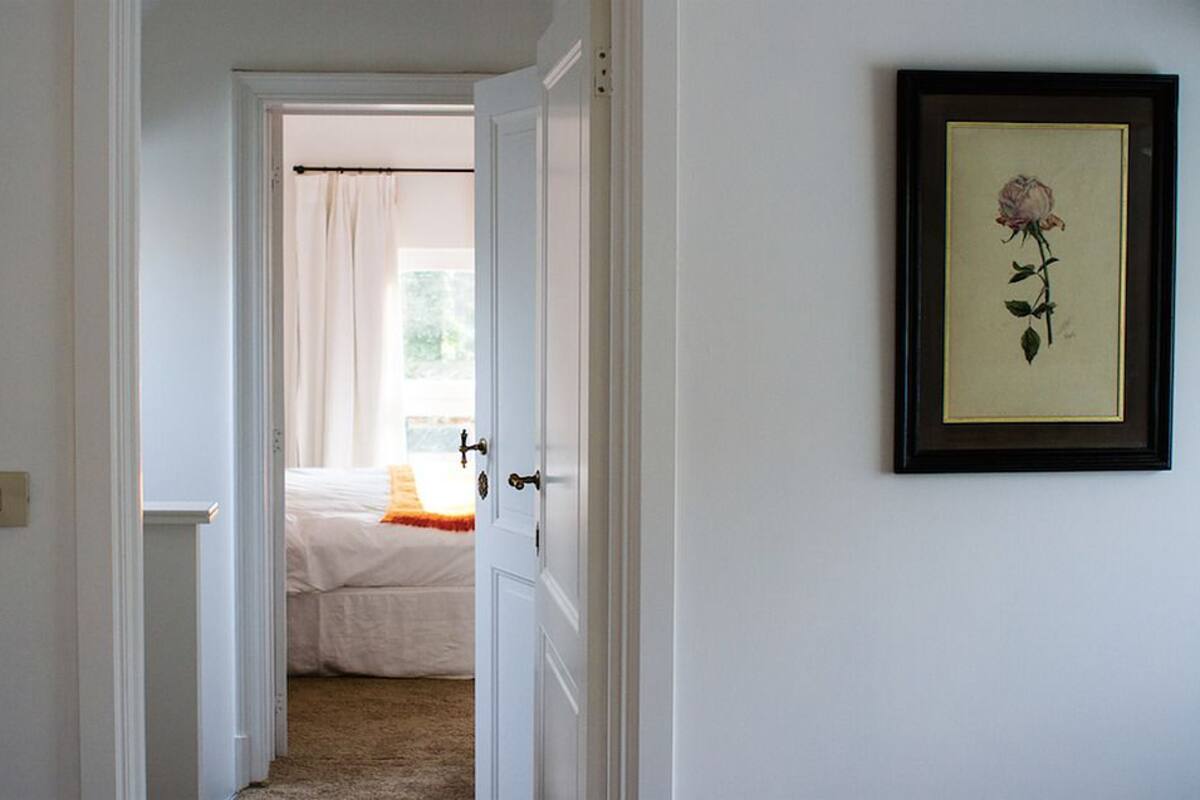 Um corredor que dá para um quarto, com uma cama, uma porta e um quadro pendurado na parede.