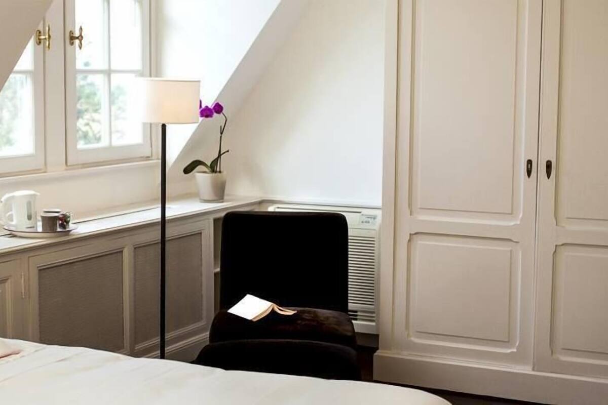 Quarto de hotel com cama, escrivaninha com abajur, planta em vaso e janela com vista.