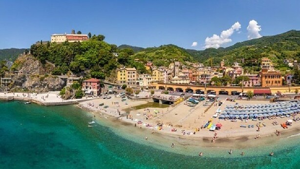 Uma vila costeira com praia, barcos e um edifício semelhante a um castelo numa colina.