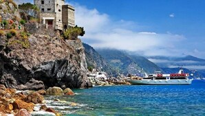 Um edifício costeiro com fachada de pedra, um barco atracado nas proximidades e montanhas ao fundo.