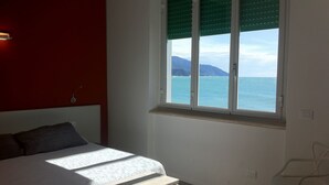 Quarto com cama, janela com persianas, uma cadeira e vista para o mar e montanhas.