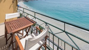 Uma varanda com mesa de madeira e cadeiras acolchoadas brancas, com vista para uma praia e águas azuis cristalinas, além de montanhas ao longe.
