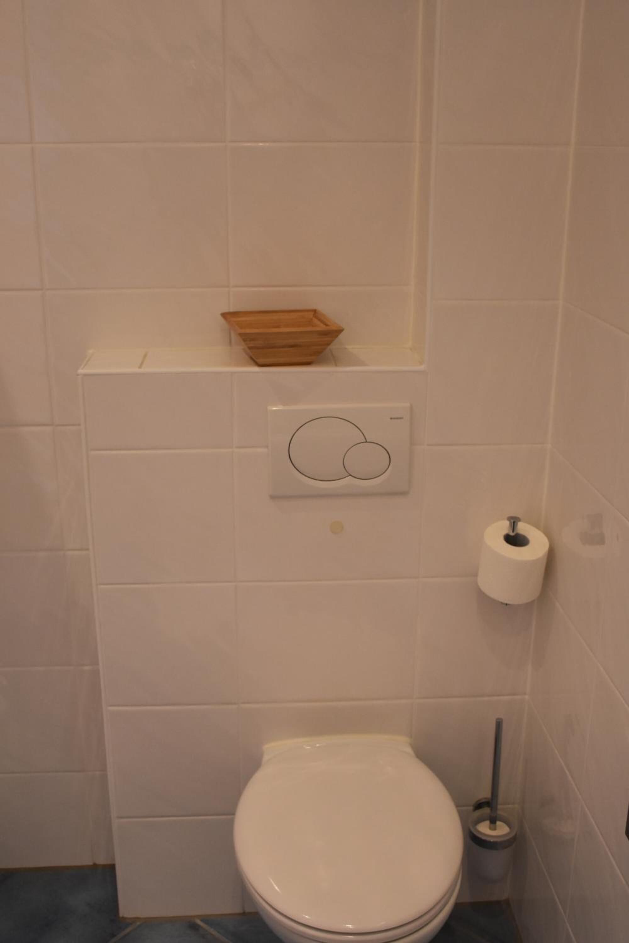 Um banheiro pequeno com vaso sanitário branco, uma prateleira de madeira acima do vaso e um rolo de papel higiênico na parede.