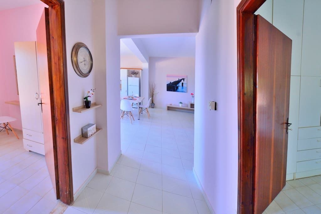 Um corredor com uma porta de madeira à direita, uma parede branca com um relógio e uma área de refeições ao fundo.