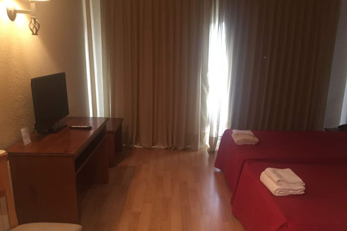 Quarto de hotel com escrivaninha de madeira, televisão, cama com cobertura vermelha e janela com cortinas.