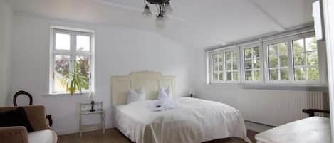 Quarto com cama grande, mesa de cabeceira, cadeira e janela com vista para árvores.