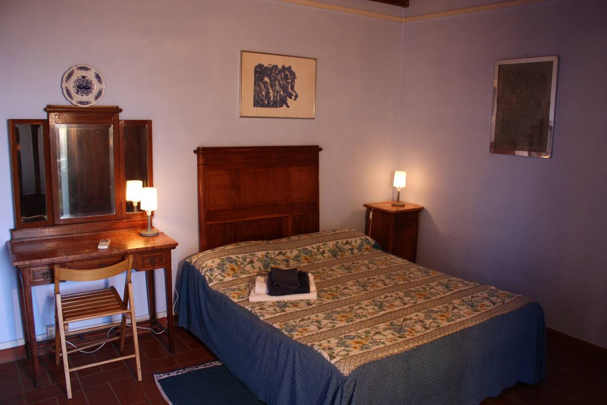 Quarto com cama de madeira, escrivaninha com cadeira, espelho e duas luminárias de cabeceira.